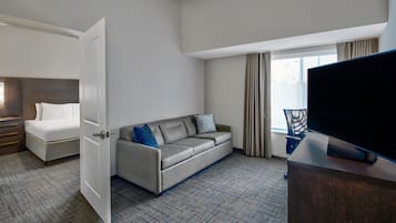 Quarto de hotel com cama, sofá, televisão e janela com cortinas.