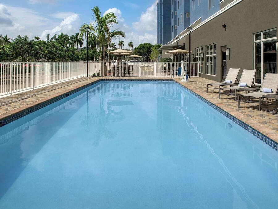 Piscina com espreguiçadeiras e guarda-sóis, cercada por um muro branco e palmeiras.