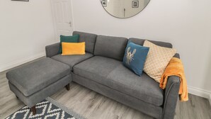 Um sofá modular cinzento com um banquinho a combinar, um espelho redondo e um tapete estampado.