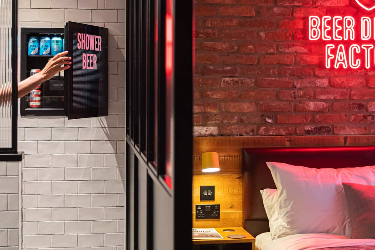 Quarto de hotel com parede de tijolos, um letreiro de neon com a inscrição 'BEER FACT' e um minibar com um aviso: 'CERVEJA DE BANHO'.