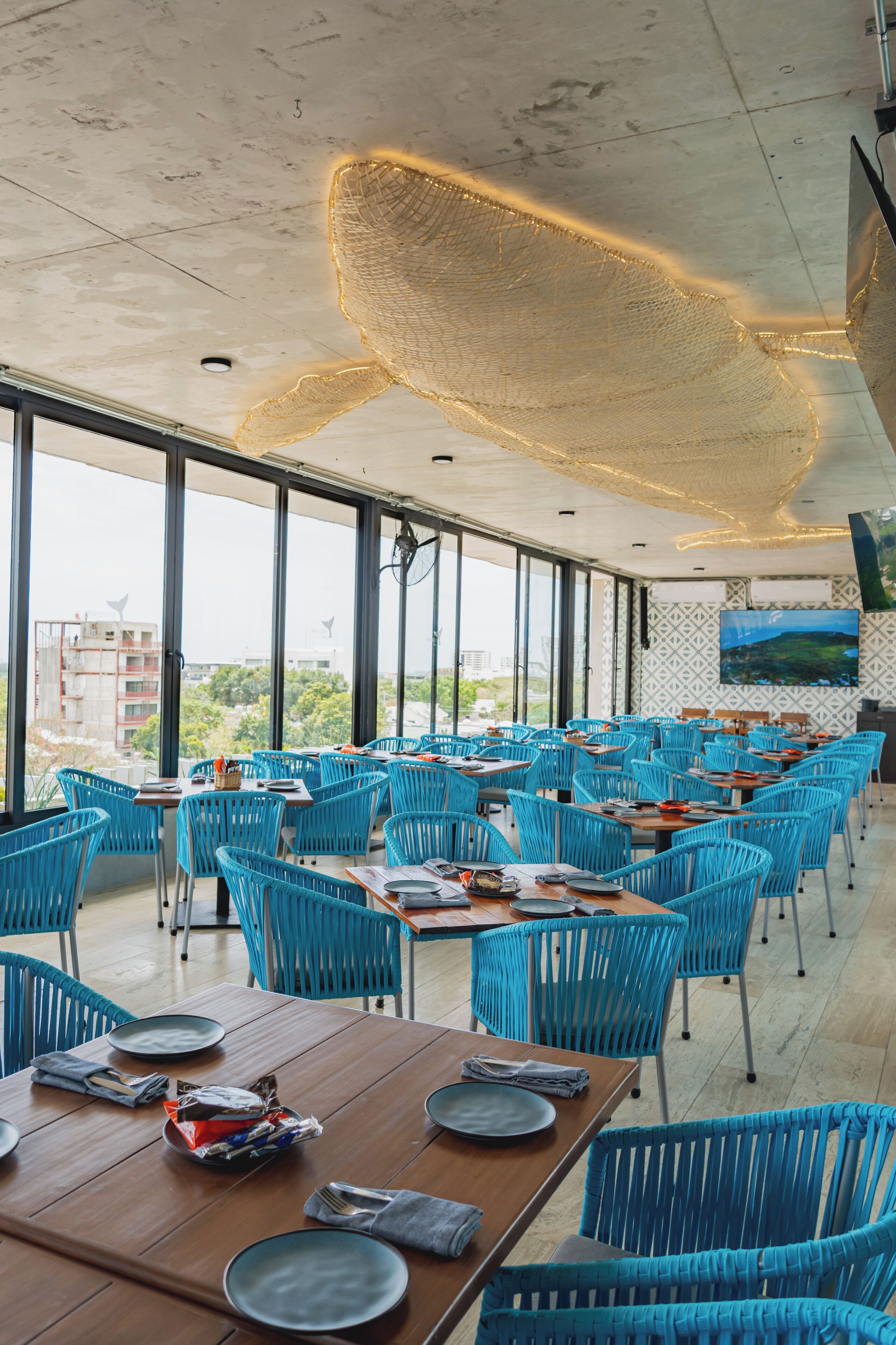 Uma sala de jantar espaçosa com cadeiras e mesas azuis, grandes janelas com vista para a cidade e um lustre.