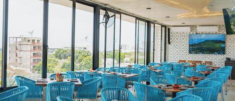Uma sala de jantar espaçosa com cadeiras e mesas azuis, grandes janelas com vista para a cidade e um lustre.