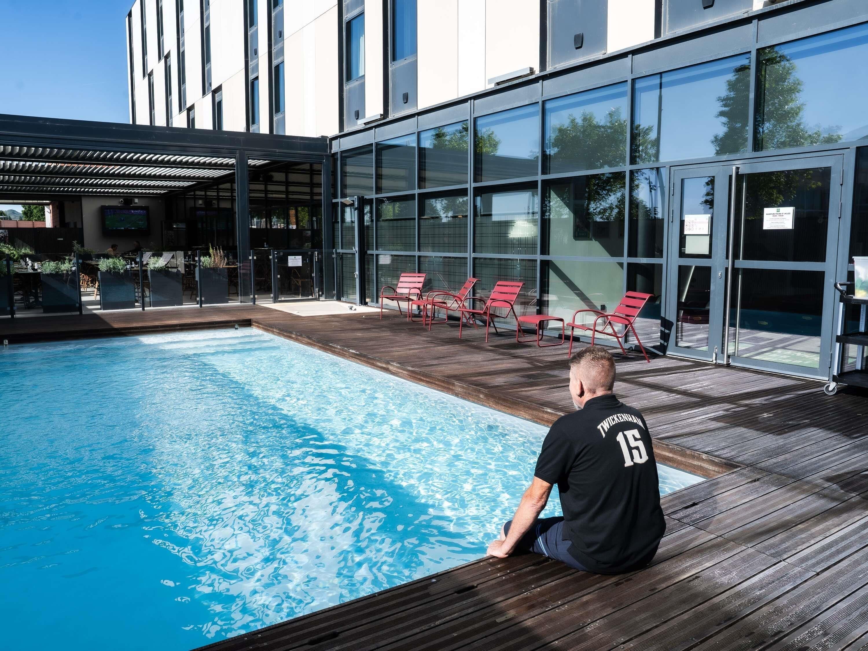 Um homem sentado à beira de uma piscina, com cadeiras vermelhas e um edifício moderno com grandes janelas ao fundo.