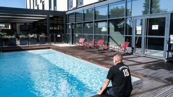 Um homem sentado à beira de uma piscina, com cadeiras vermelhas e um edifício moderno com grandes janelas ao fundo.