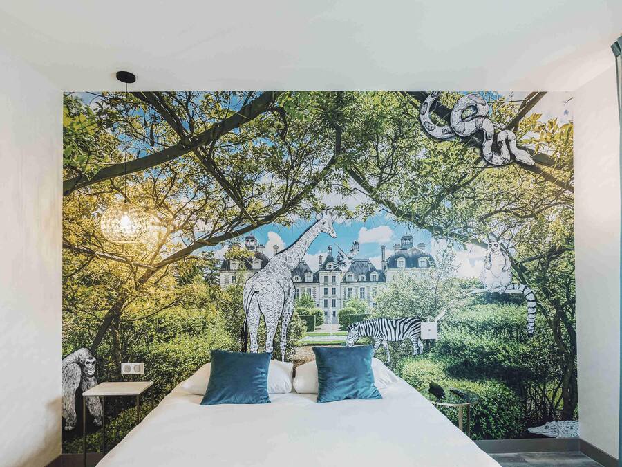Quarto com mural de uma floresta e animais, cama com almofadas azuis e vista de um edifício ao fundo.