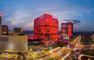 Hotel moderno com fachada vermelha marcante, entrada bem iluminada e vista do horizonte urbano.
