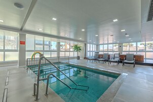 Piscina interior com paredes de vidro transparente, escada de metal e várias espreguiçadeiras.