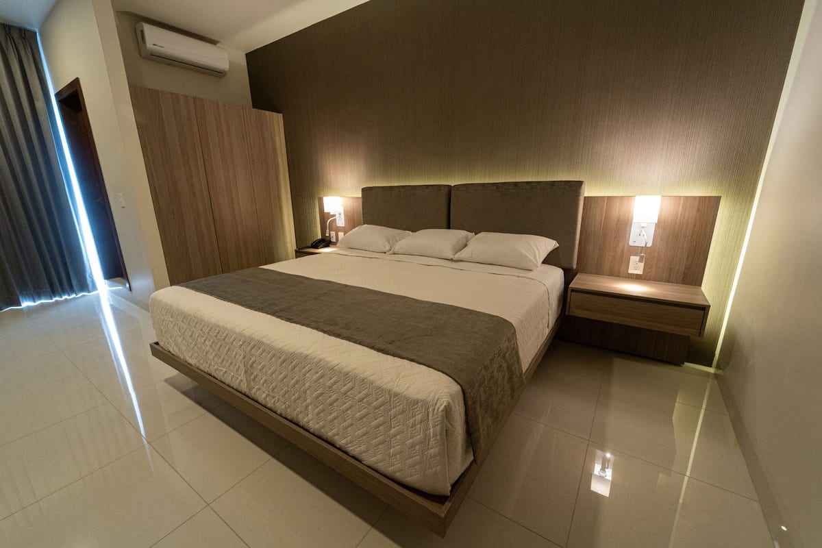 Quarto de hotel moderno com cama grande, cabeceira de madeira, mesas de cabeceira com luminárias e vista para o exterior.
