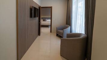 Quarto de hotel moderno com televisão de tela plana na parede, poltrona confortável, cama com cabeceira e uma janela grande com cortinas.