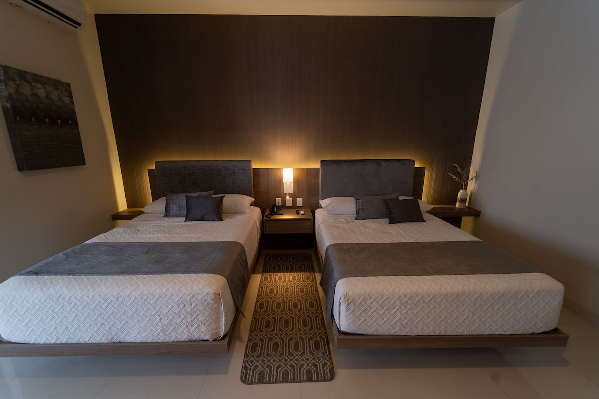 Quarto de hotel moderno com duas camas, uma mesinha de cabeceira, um abajur e um tapete decorativo.
