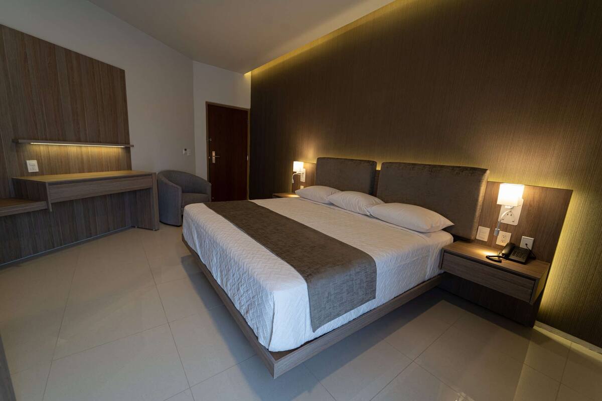 Quarto de hotel moderno com uma cama grande, mesas de cabeceira e uma escrivaninha embutida.