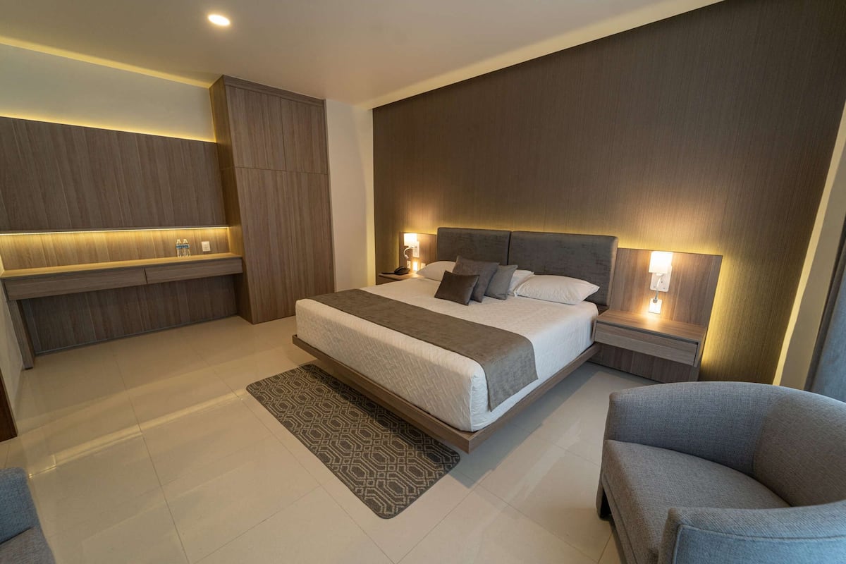 Quarto moderno com cama grande, mesas de cabeceira com luminárias, roupeiro embutido e um pequeno espaço de estar.