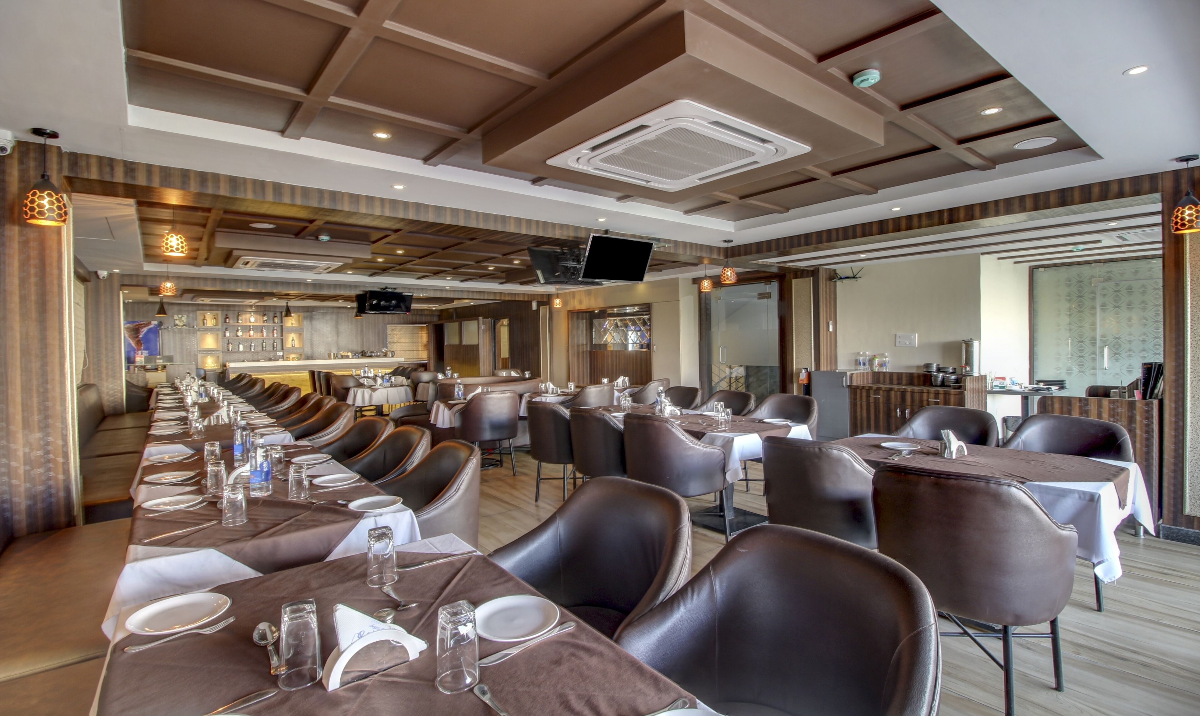 Um restaurante com mesas grandes e partilhadas, e também cabines individuais, com talheres e um ecrã de televisão.