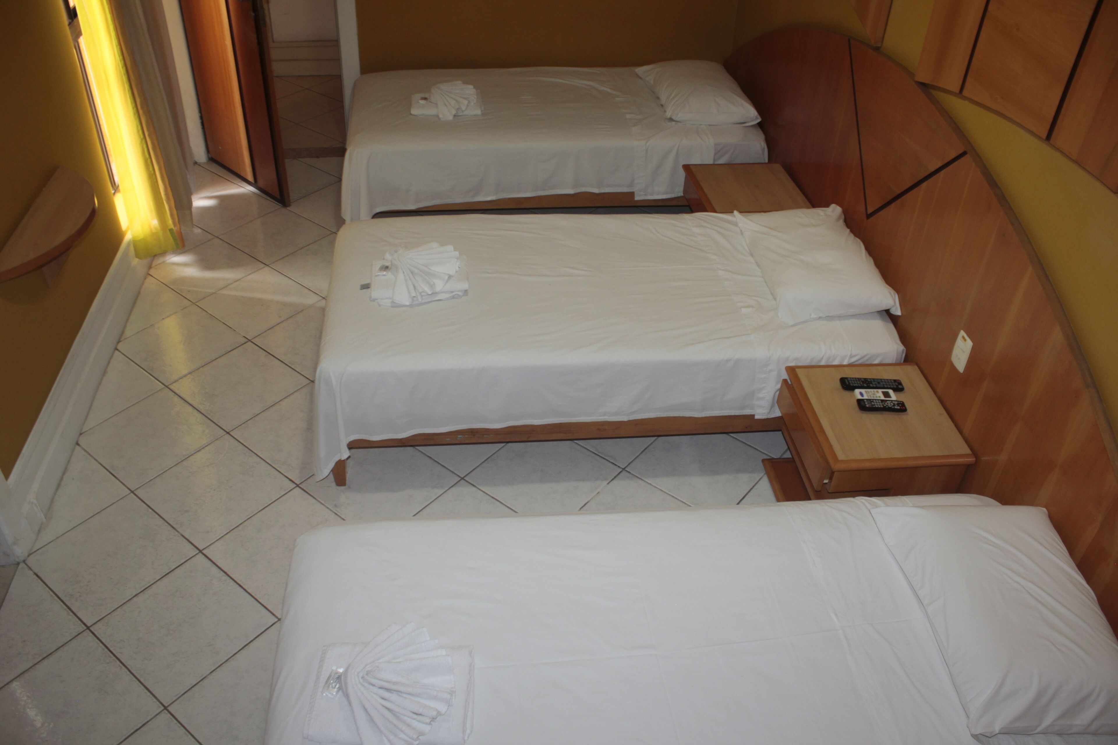 Quarto de hotel com duas camas de solteiro, cabeceira de madeira, mesa de cabeceira de madeira pequena com controle remoto e piso de azulejo.