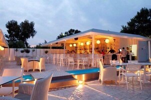 Uma área de refeições ao ar livre com mesas e cadeiras brancas, uma piscina e um espaço de estar coberto com luminárias pendentes.