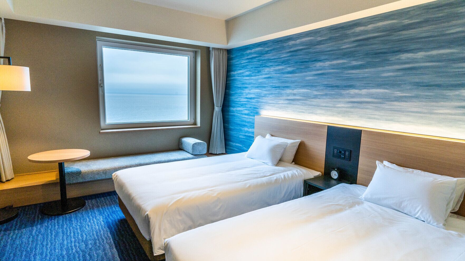 Quarto de hotel com duas camas, uma mesa pequena, uma luminária, uma janela com vista para o mar e uma parede com um desenho abstrato azul.