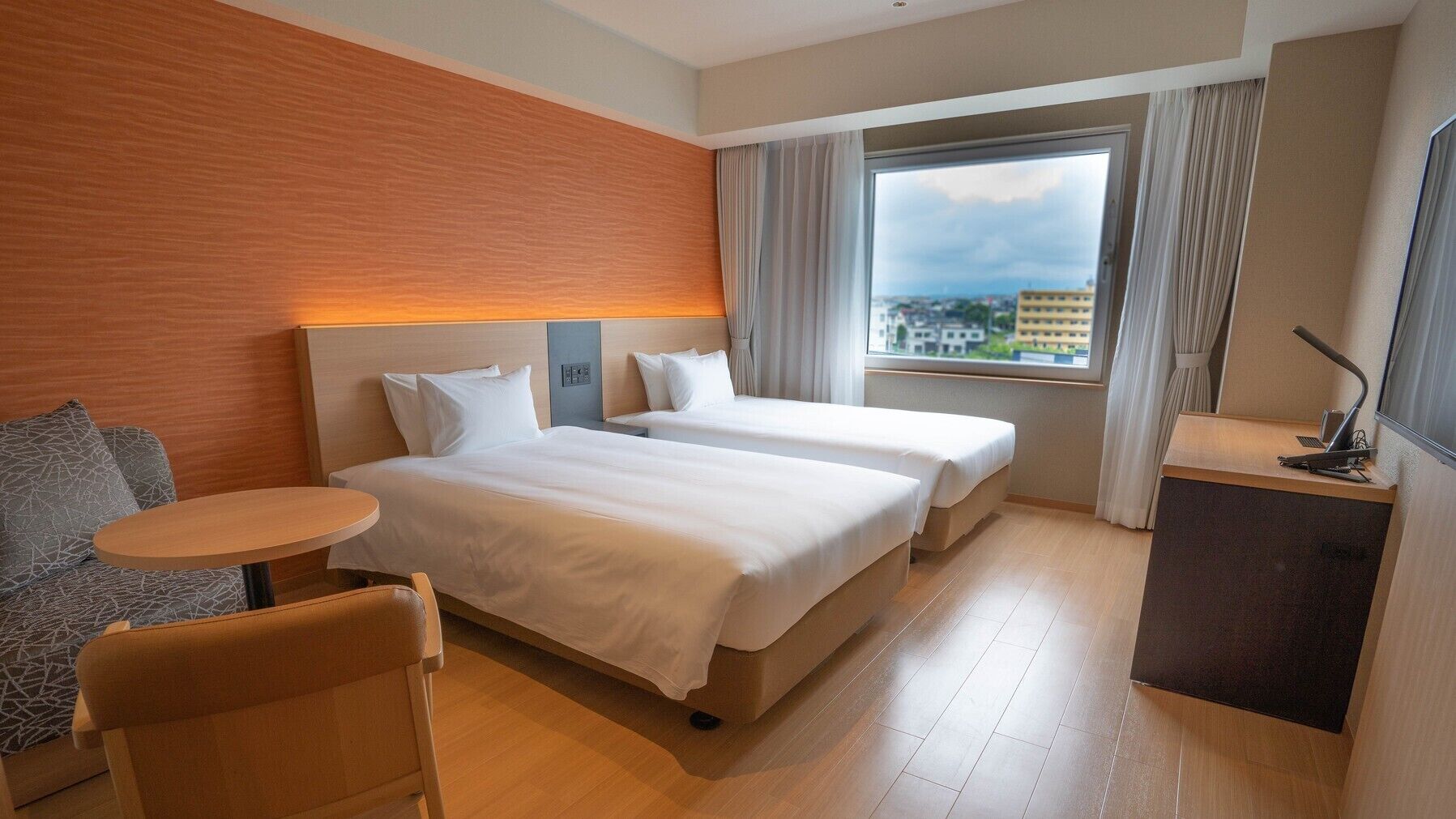 Quarto de hotel com duas camas, uma secretária pequena, uma cadeira e vista para a cidade.