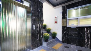 Elevador com visor digital, parede de mármore, escada com piso tátil e um sinal de informações.