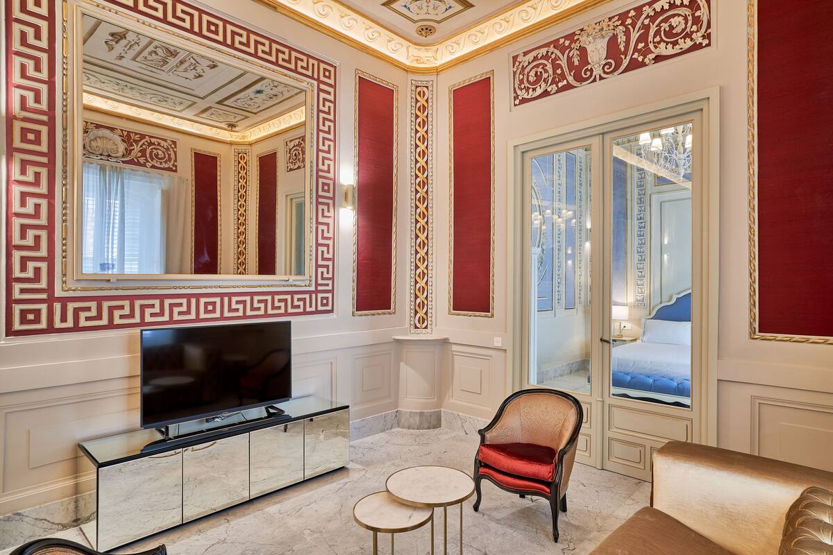Quarto luxuoso com um espelho grande, televisão, poltrona vermelha estofada e uma cama com cabeceira azul.