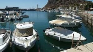 Marina com vários barcos atracados, um muro de pedra e uma colina com edifícios ao fundo.