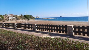 Uma praia com um calçadão pavimentado, uma cerca de madeira e um afloramento rochoso ao longe.