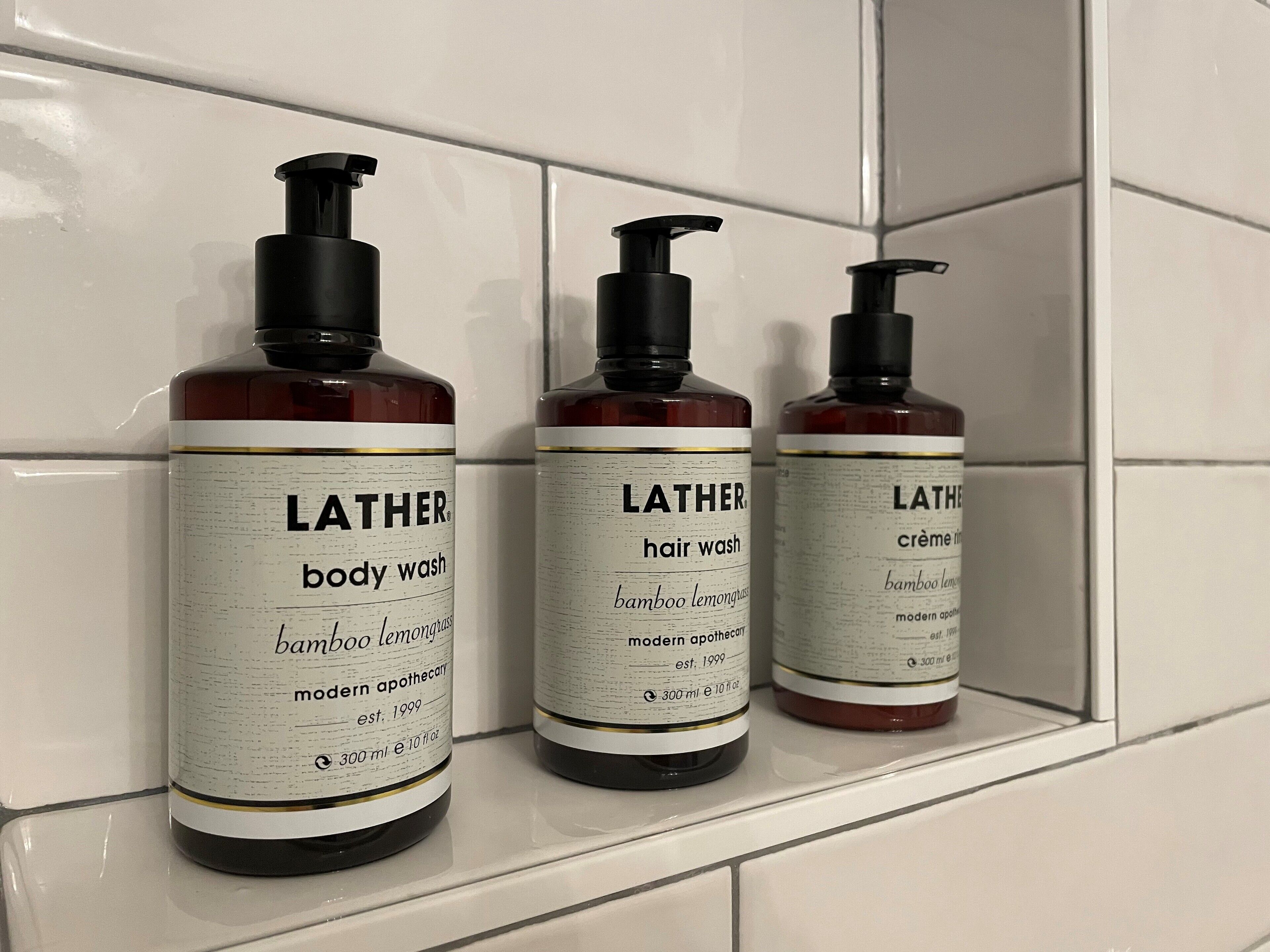 Três embalagens de Lather, incluindo sabonete líquido, shampoo e creme, sobre uma prateleira revestida com azulejos.