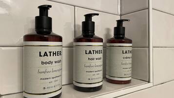 Três embalagens de Lather, incluindo sabonete líquido, shampoo e creme, sobre uma prateleira revestida com azulejos.