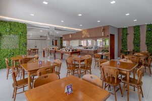 Um café moderno com mesas e cadeiras de madeira, uma parede verde com um letreiro e uma área de cozinha ao fundo.