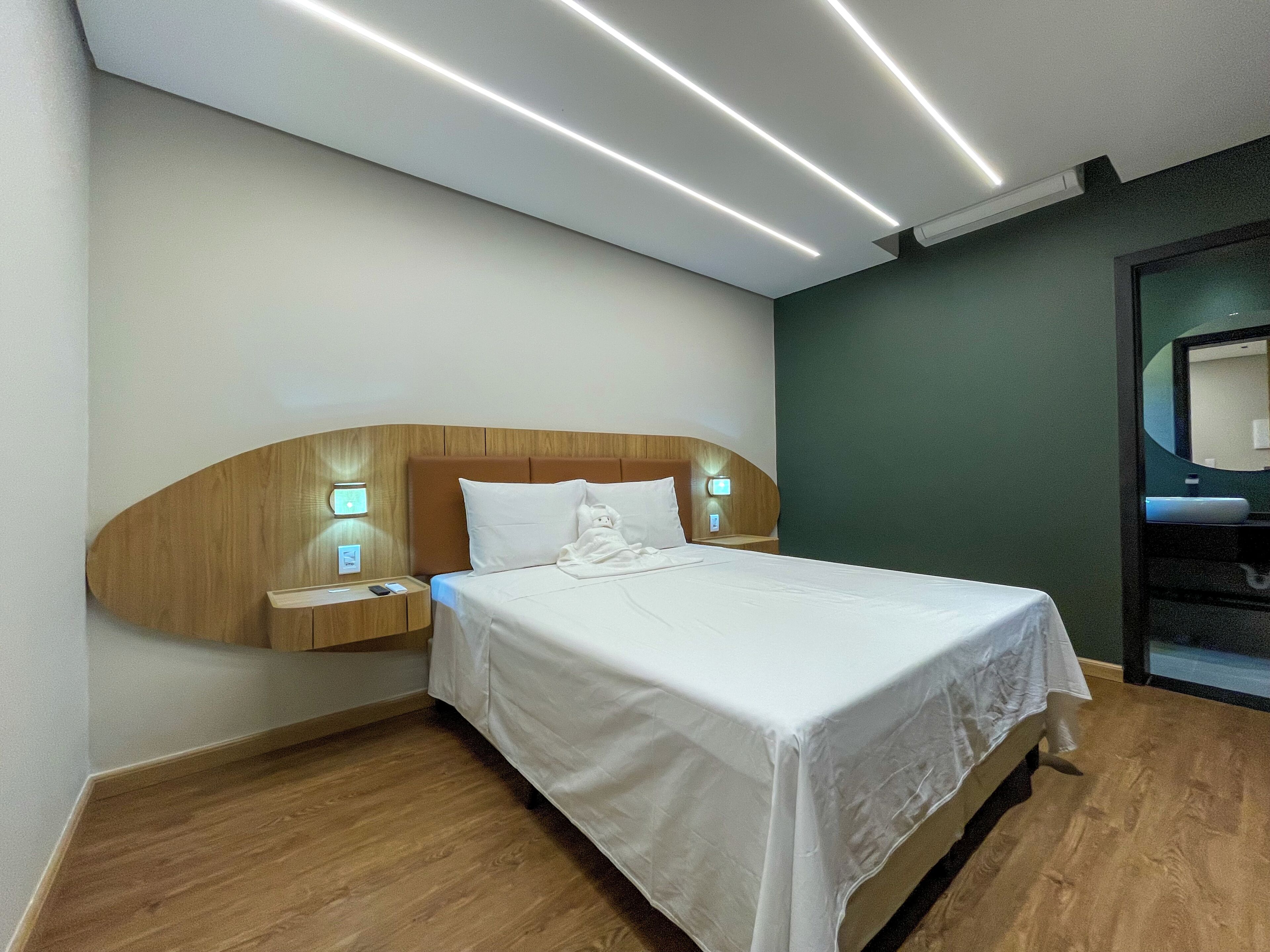 Quarto de hotel com cabeceira de madeira, cama com roupa de cama branca, mesa de cabeceira com abajur e espelho na parede.