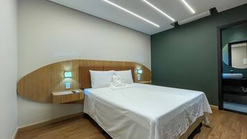 Quarto de hotel com cabeceira de madeira, cama com roupa de cama branca, mesa de cabeceira com abajur e espelho na parede.