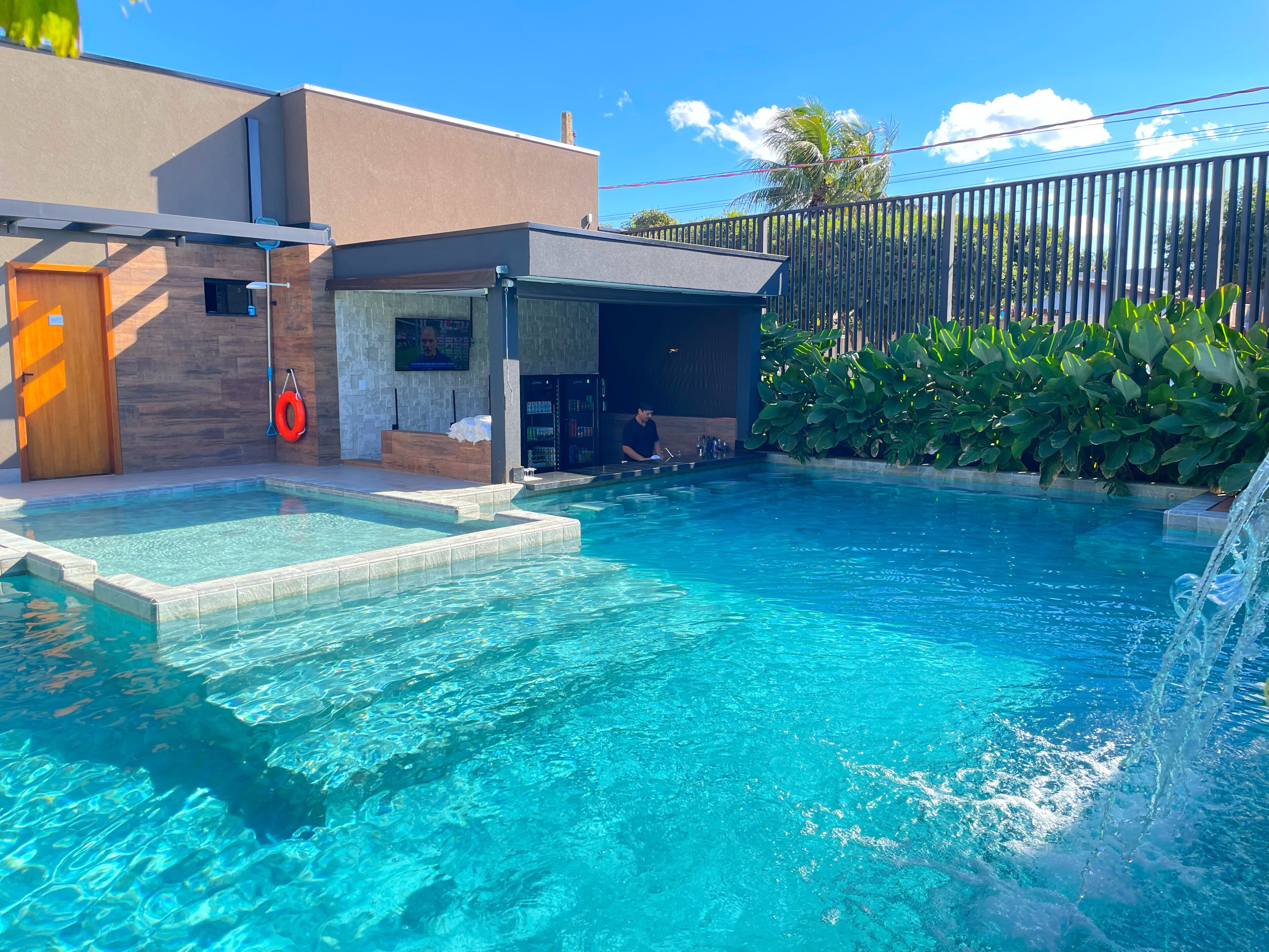Piscina com área de bar, boia salva-vidas e um edifício moderno com porta de madeira.
