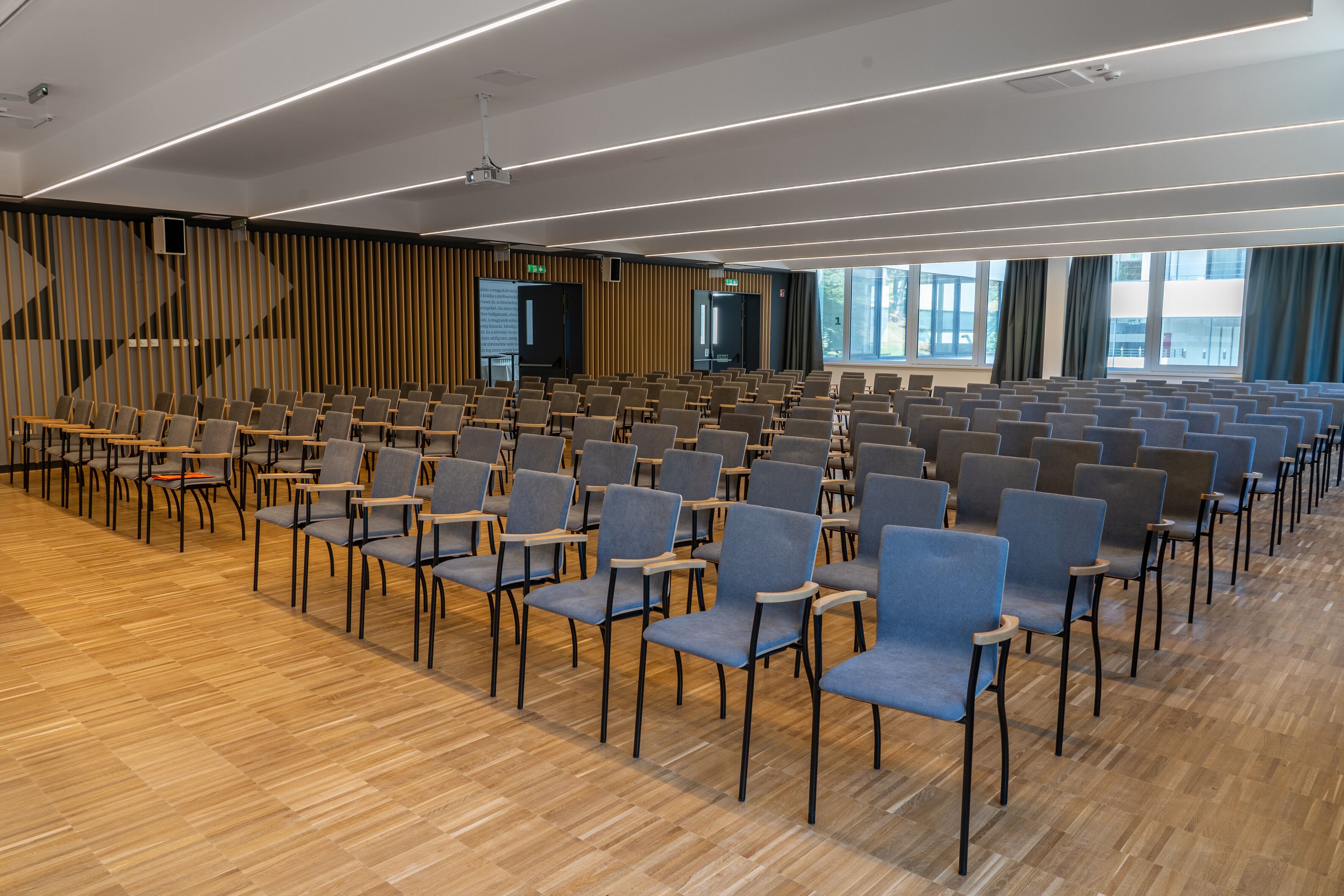 Uma sala de conferências com fileiras de cadeiras, um projetor e grandes janelas.