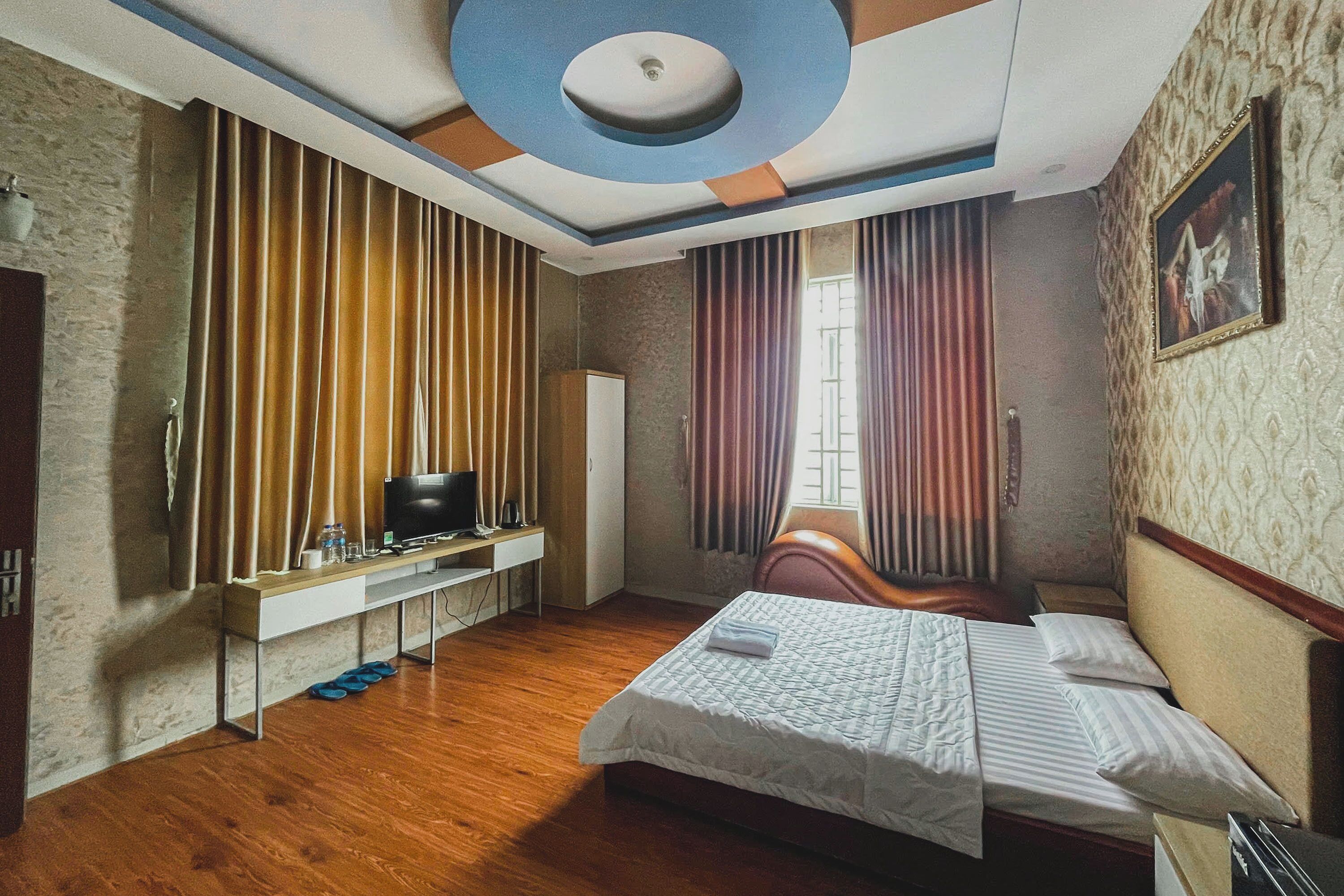 Quarto de hotel com cama, televisão em suporte, guarda-roupa e janela com cortinas.