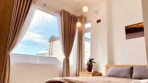 Quarto de hotel com uma janela grande, cama com cabeceira de madeira, mesa de cabeceira com uma planta e um quadro na parede.