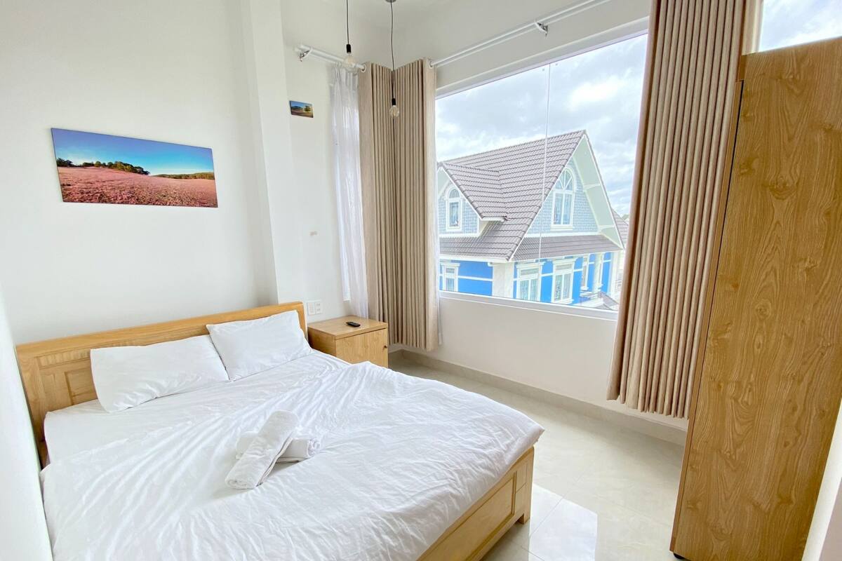 Um quarto com uma cama grande, uma mesa de cabeceira, uma janela com cortinas e vista para uma casa.