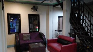 Sala de estar com um sofá roxo, um sofá vermelho e uma mesa de centro de vidro. Há um ventilador de teto e uma escada com corrimão de metal.
