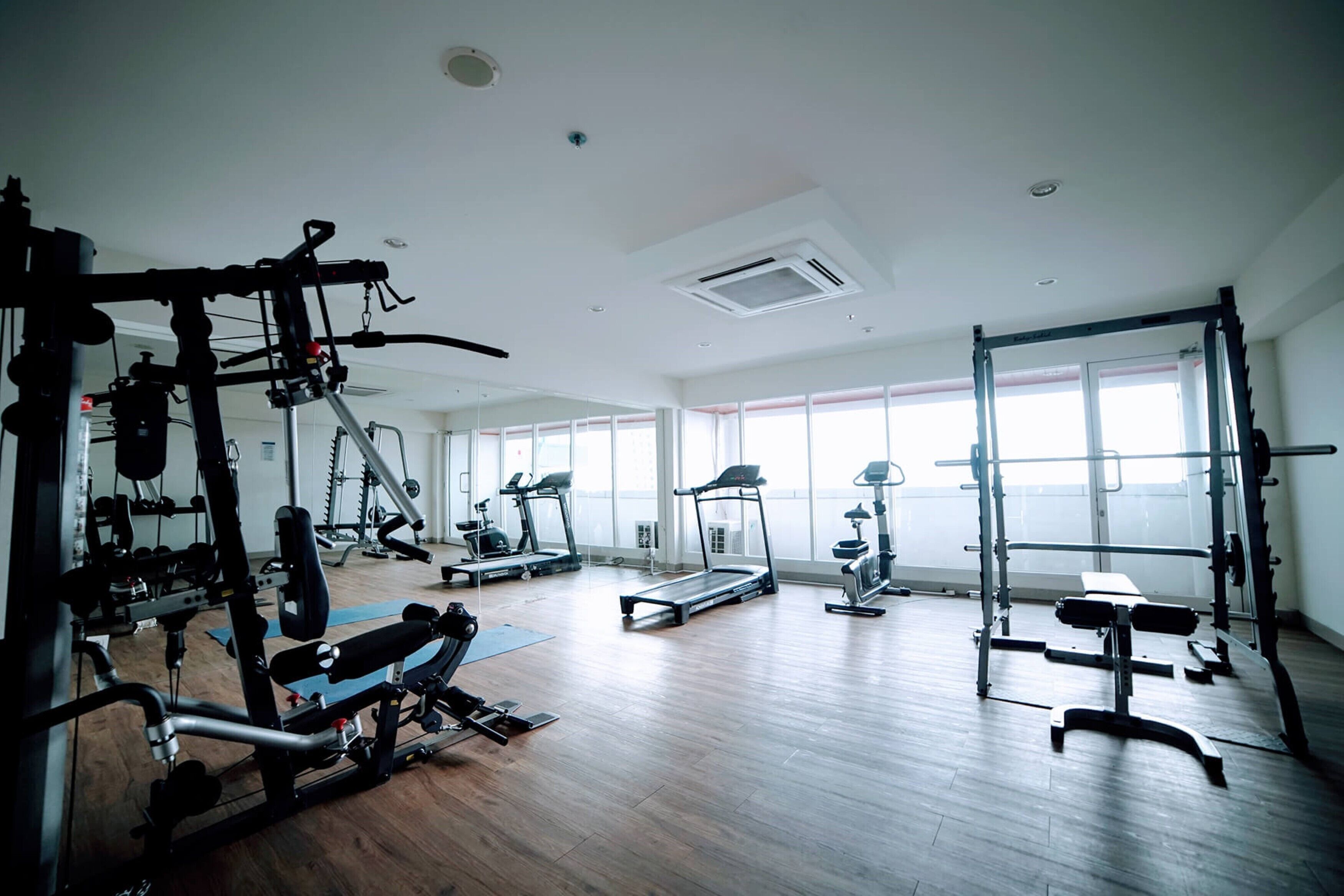 Uma academia ampla com diversos equipamentos de exercício, amplas janelas e piso de madeira.