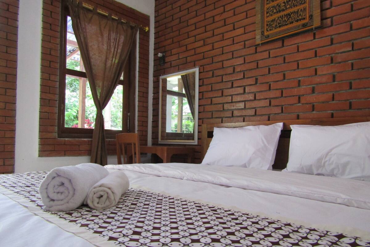 Quarto de hotel com parede de tijolos, cama com roupa de cama branca, colcha estampada e janela com cortinas.