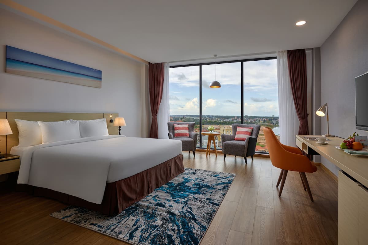 Quarto de hotel com uma cama grande, uma escrivaninha, duas cadeiras, vista para a cidade e um quadro na parede.