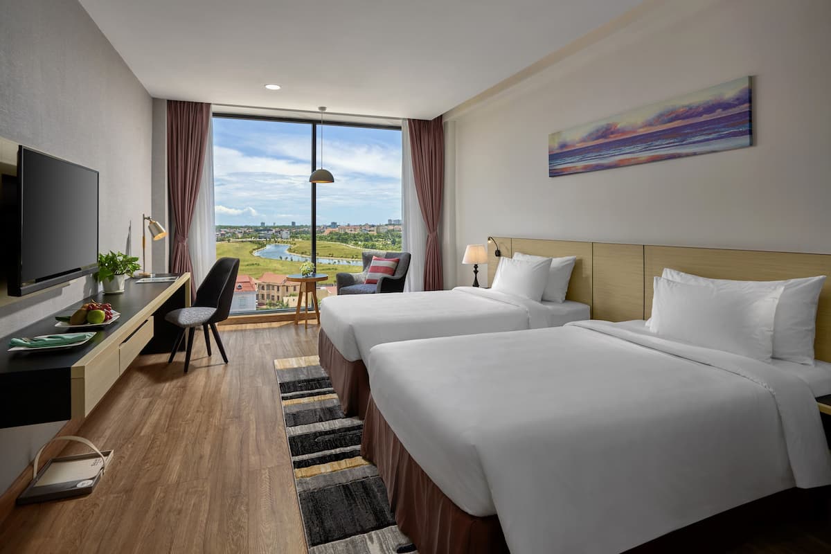 Quarto de hotel com duas camas, uma mesa de trabalho, televisão e uma janela ampla com vista para a água e o horizonte urbano.