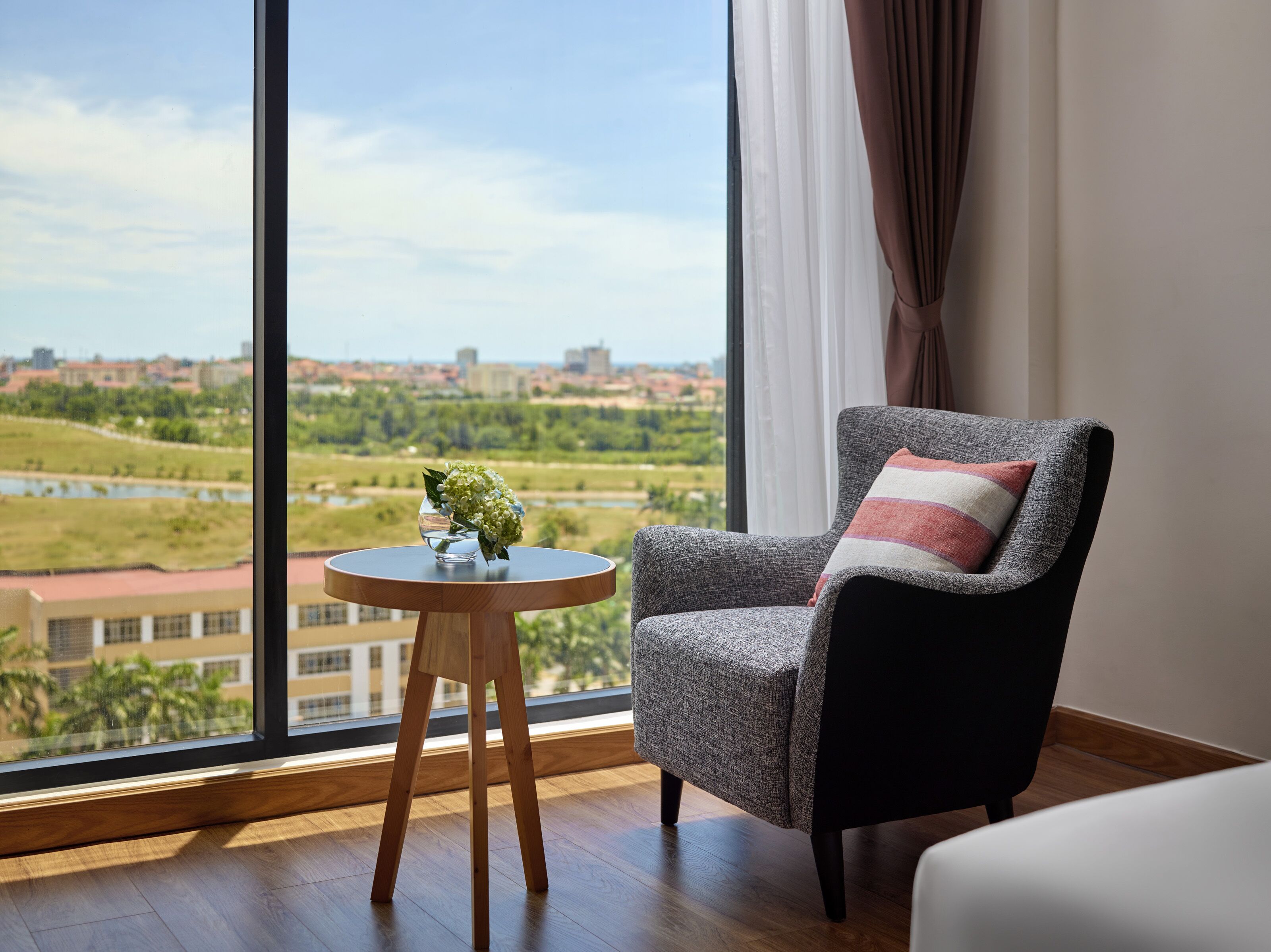 Quarto com uma janela grande, uma mesa de madeira redonda com um vaso de flores e uma poltrona cinza com almofada listrada.
