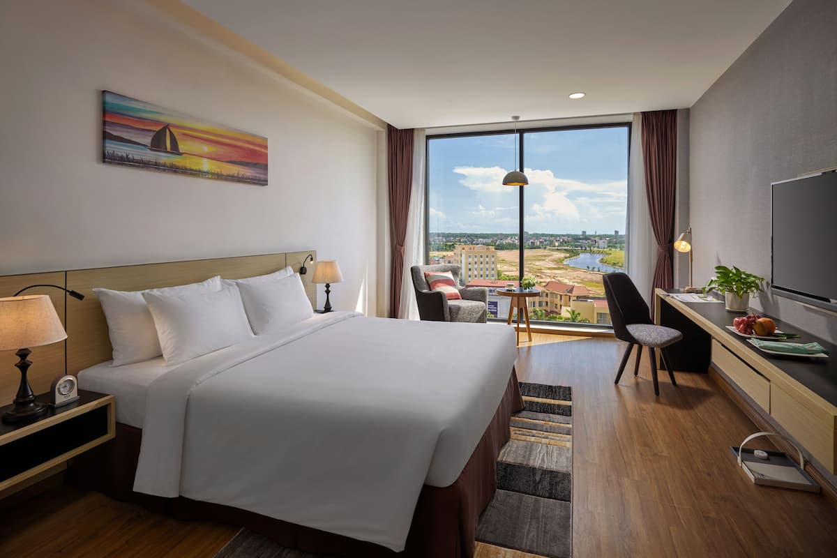 Quarto de hotel com cama, escrivaninha, cadeira, televisão e uma janela grande com vista para a cidade.