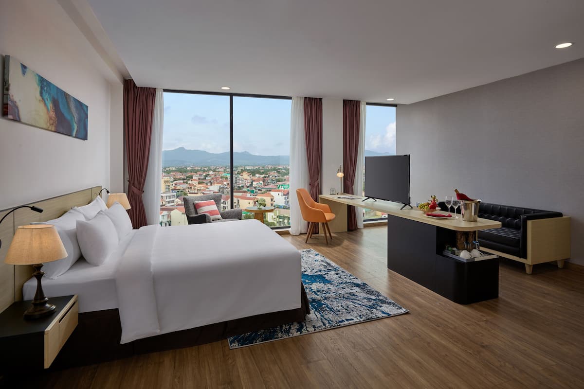 Quarto de hotel com cama, escrivaninha e sofá, janelas amplas com vista para a cidade e um quadro na parede.