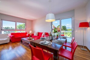 Sala de estar moderna com sofá vermelho, mesa de jantar para quatro pessoas e grandes janelas com vista para o verde.
