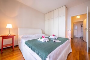 Uma cama bem arrumada com um cobertor verde, uma mesinha de cabeceira vermelha com uma luminária, e um guarda-roupa branco.