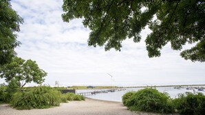 Uma praia arenosa com um píer que se estende por águas calmas, cercada por vegetação e turbinas eólicas ao longe.