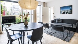 Uma sala de estar moderna com uma mesa de jantar redonda, cadeiras pretas, um sofá cinzento e uma grande janela com vista para a cidade.