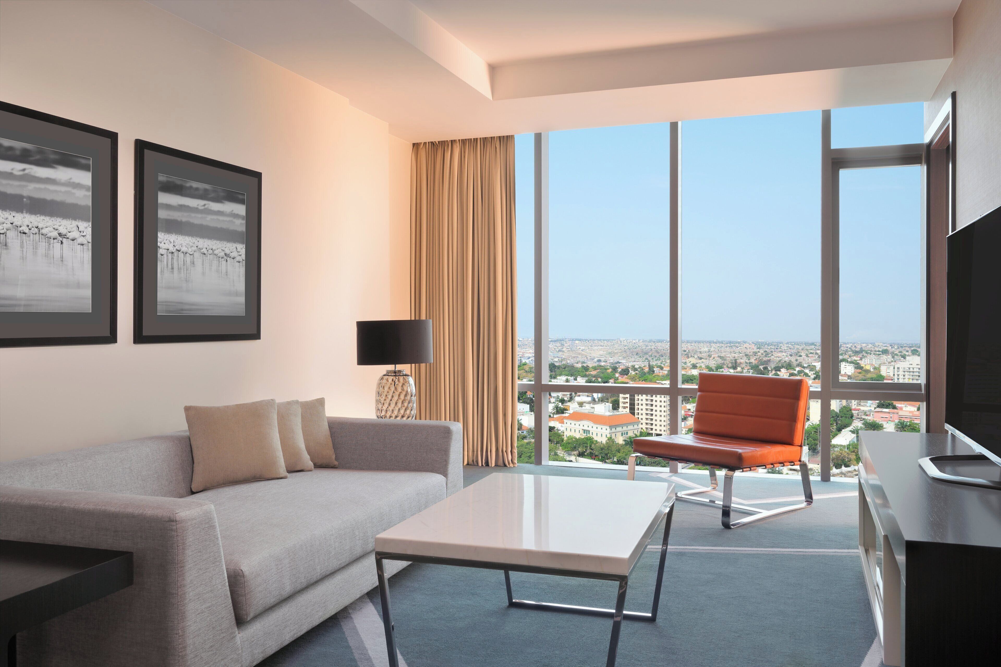 Quarto de hotel moderno com uma grande janela que oferece vista para a cidade, um sofá cinza, uma mesa pequena e uma televisão.