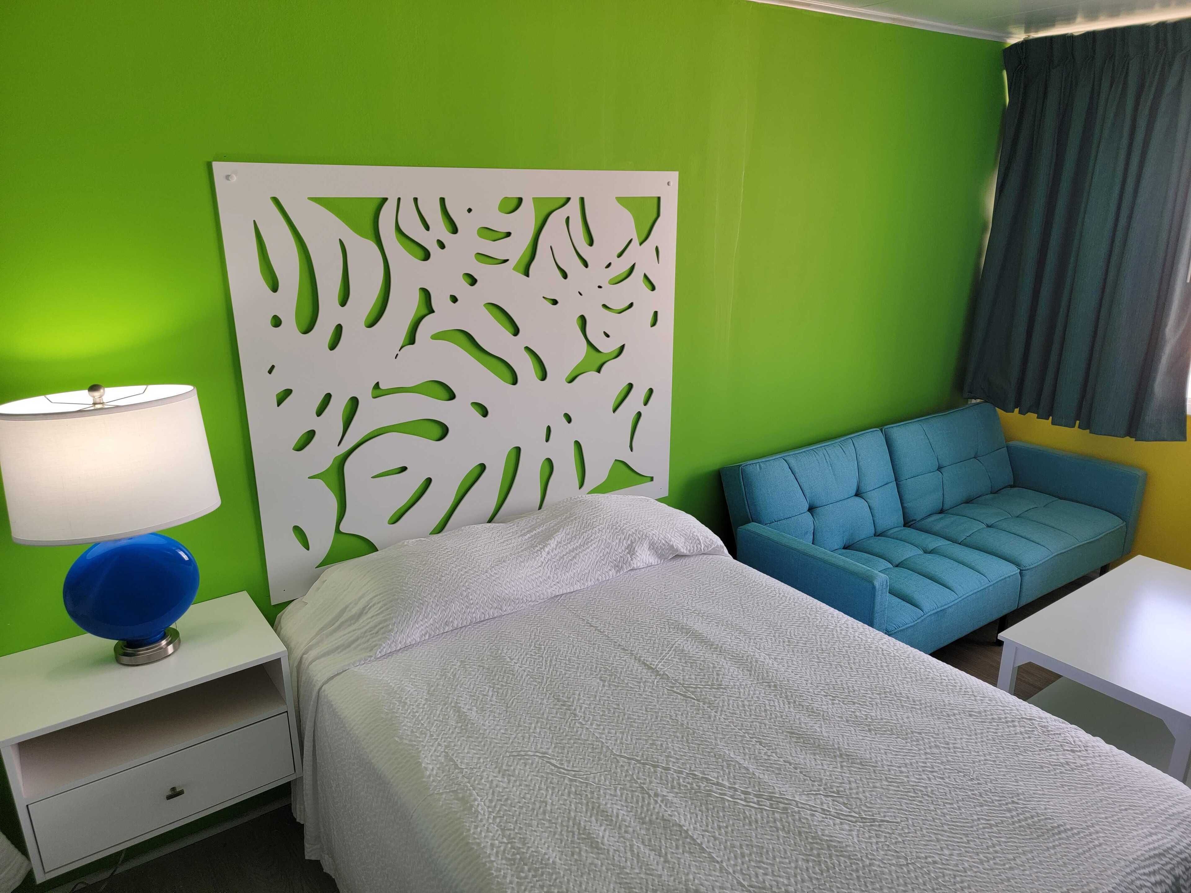 Quarto com uma cama, um sofá azul, uma mesa de cabeceira branca com abajur e uma parede verde com painel decorativo.