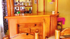 Uma área de bar com balcão de madeira, prateleiras cheias de garrafas de bebidas, um espelho e toalha de mesa rosa.